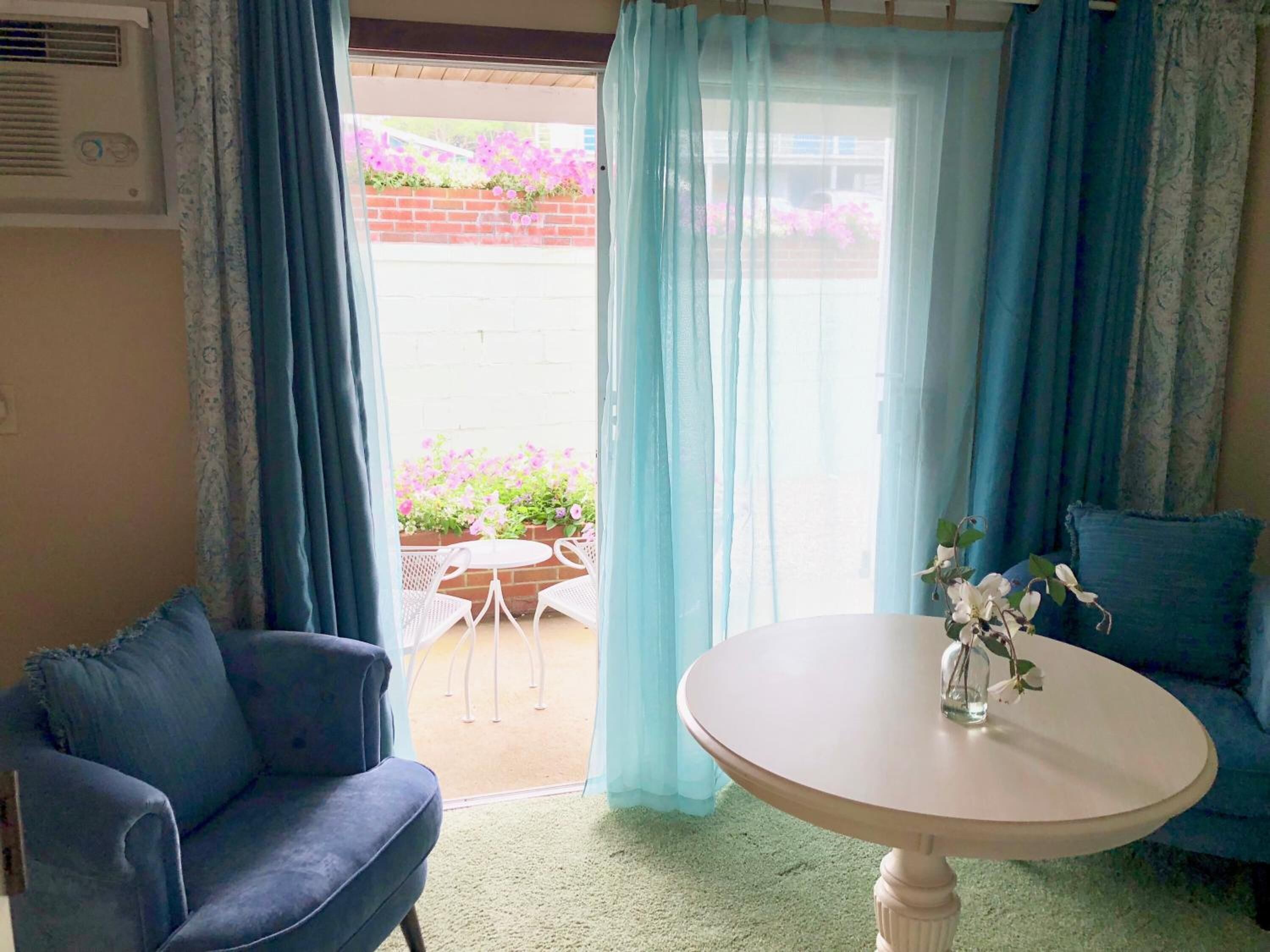 Quarto com sofá azul, mesa redonda com um vaso de flores e vista para um pátio com parede de tijolos e flores cor-de-rosa.