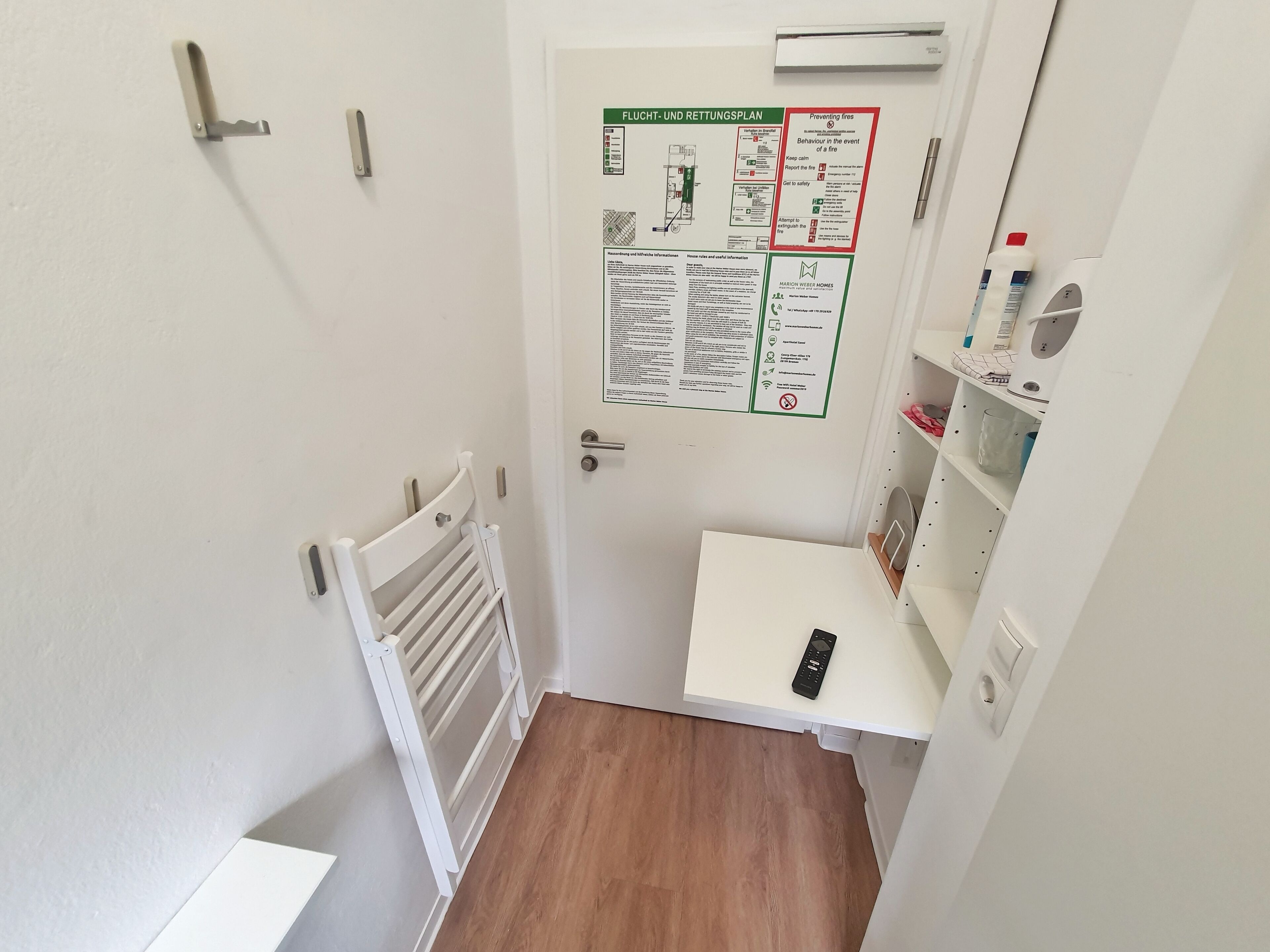 Quarto pequeno e limpo com porta branca, radiador, prateleira com controle remoto e ar condicionado de parede.