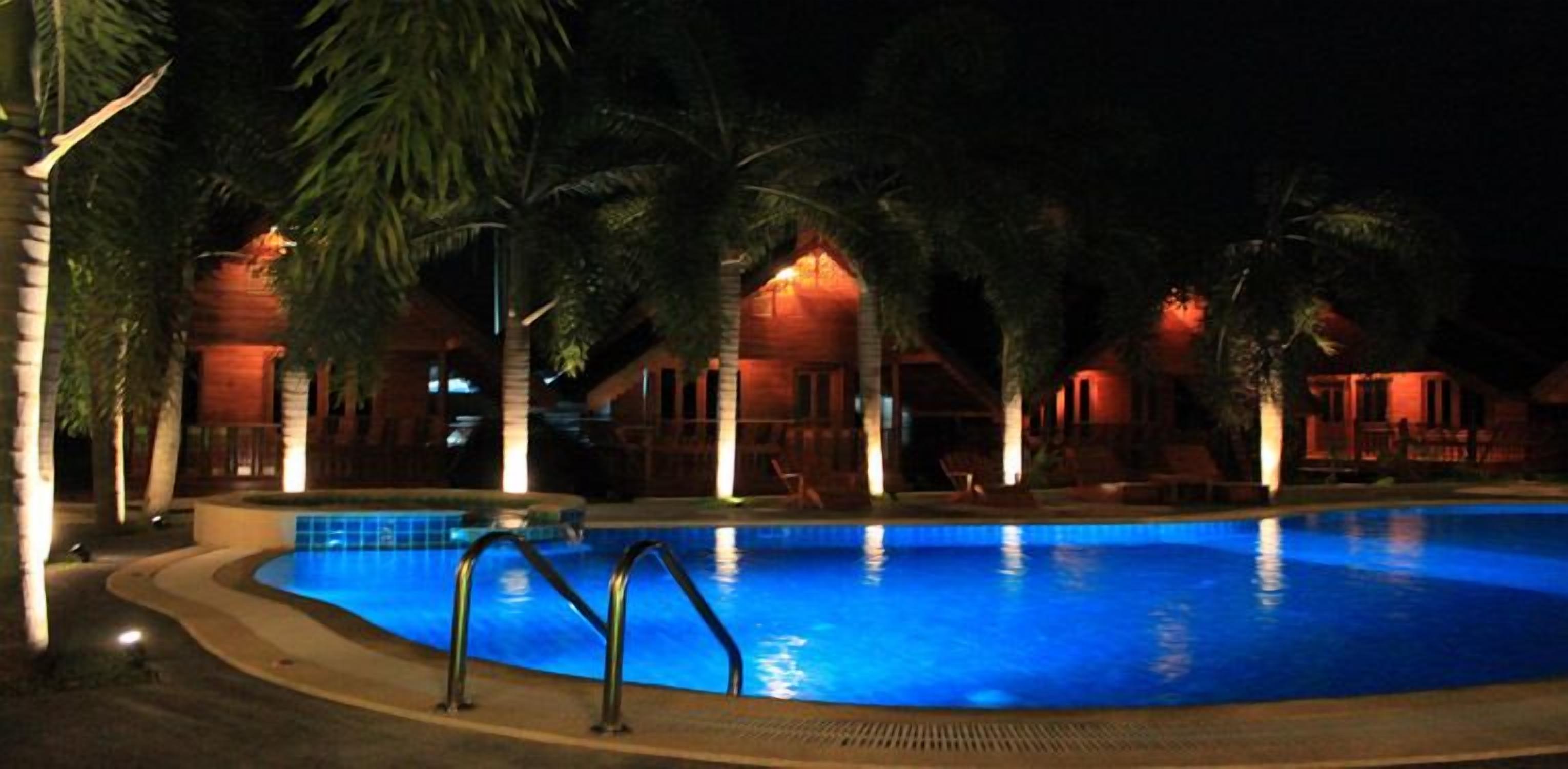 Piscina à noite com palmeiras e um edifício ao fundo.