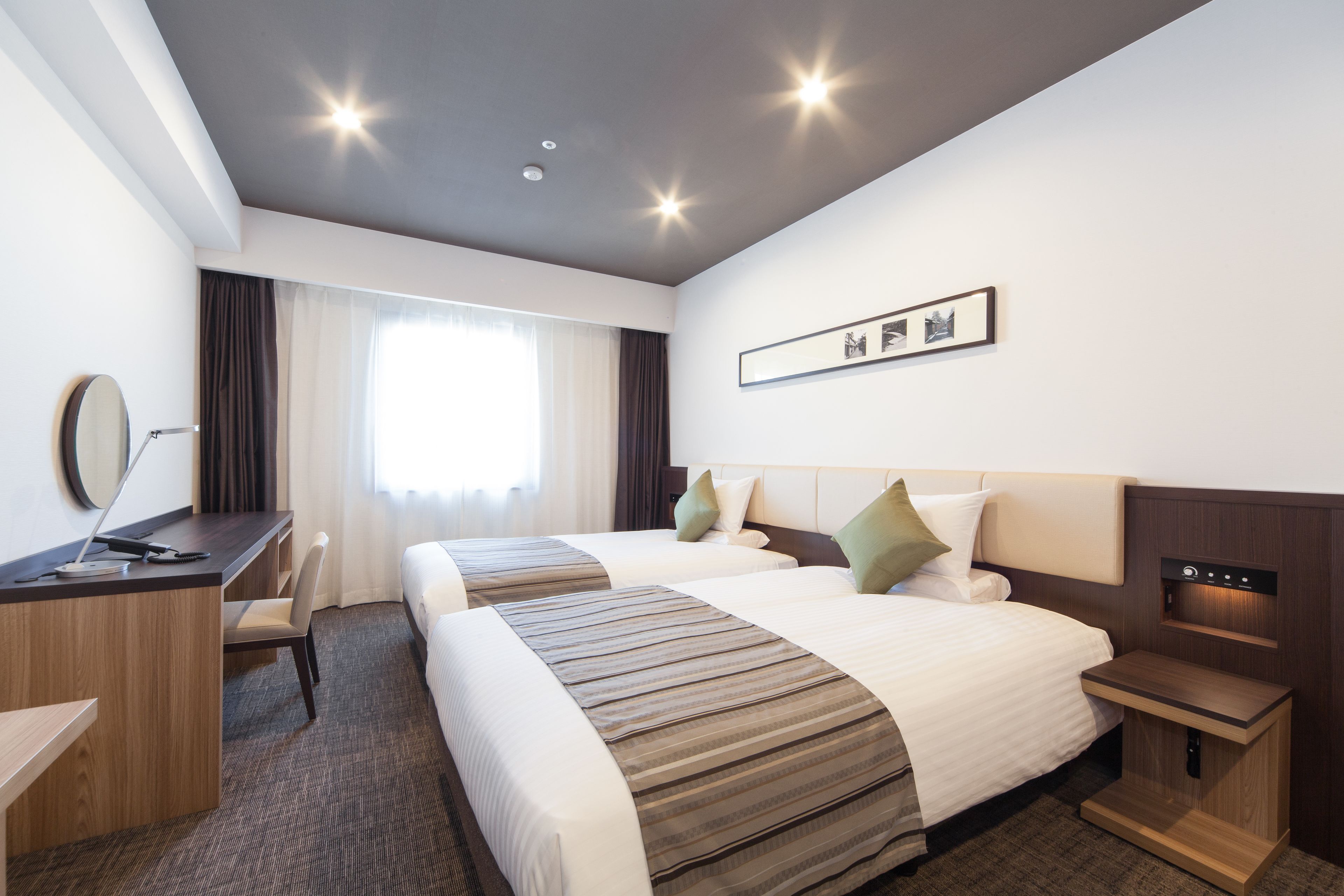 Quarto de hotel com duas camas, uma escrivaninha, uma cadeira, um espelho, mesa de cabeceira e uma obra de arte na parede.