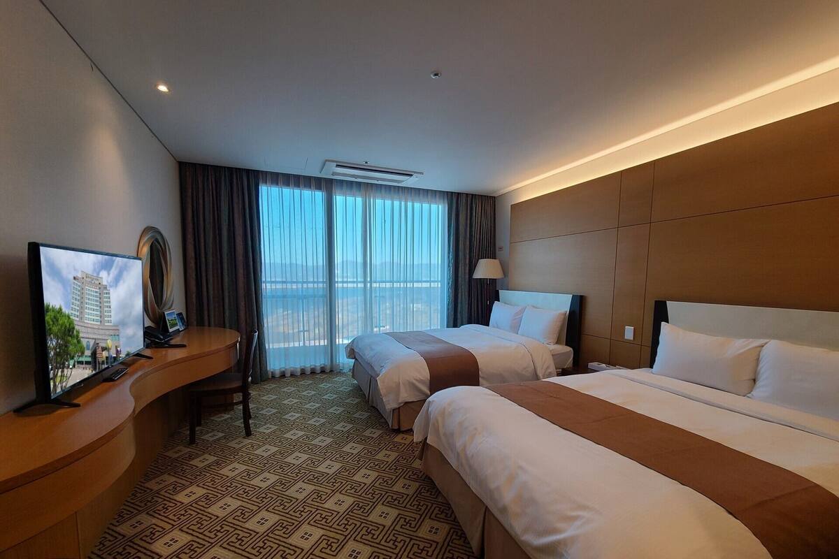 Quarto de hotel com duas camas, uma mesa de trabalho com televisão e vista para a cidade através de amplas janelas.