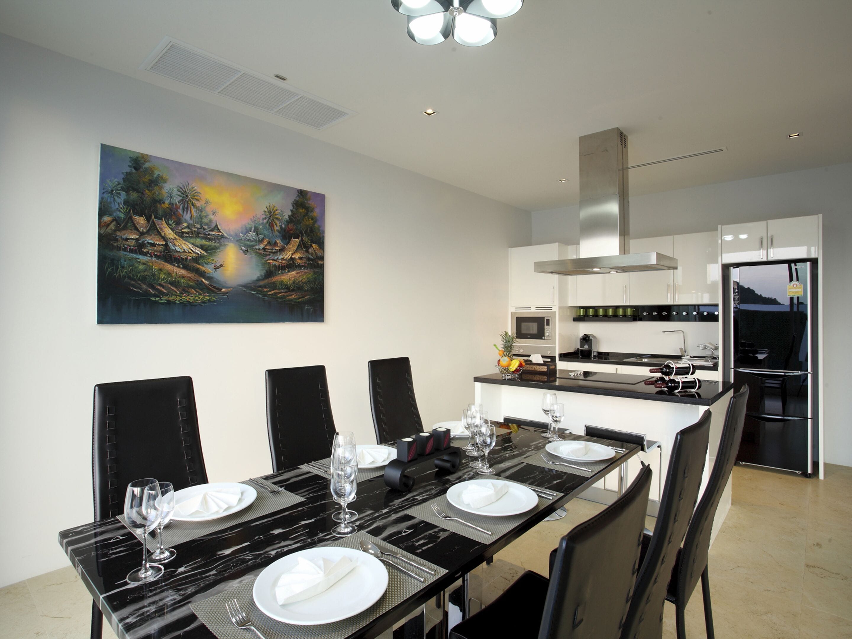 Uma sala de jantar moderna com uma mesa comprida posta para quatro pessoas, cozinha ao fundo e um quadro na parede.