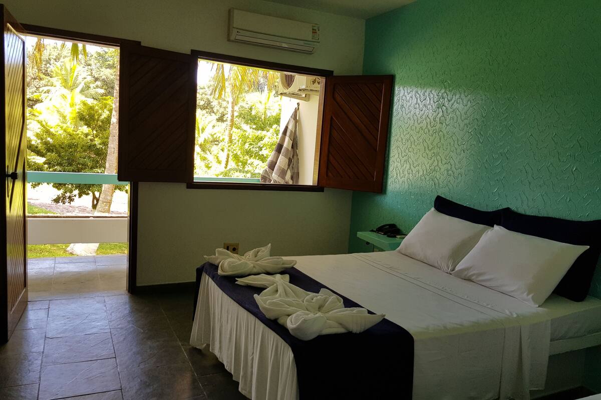 Quarto de hotel com cama, mesa de cabeceira, janela com cortinas e vista para o verde lá fora.