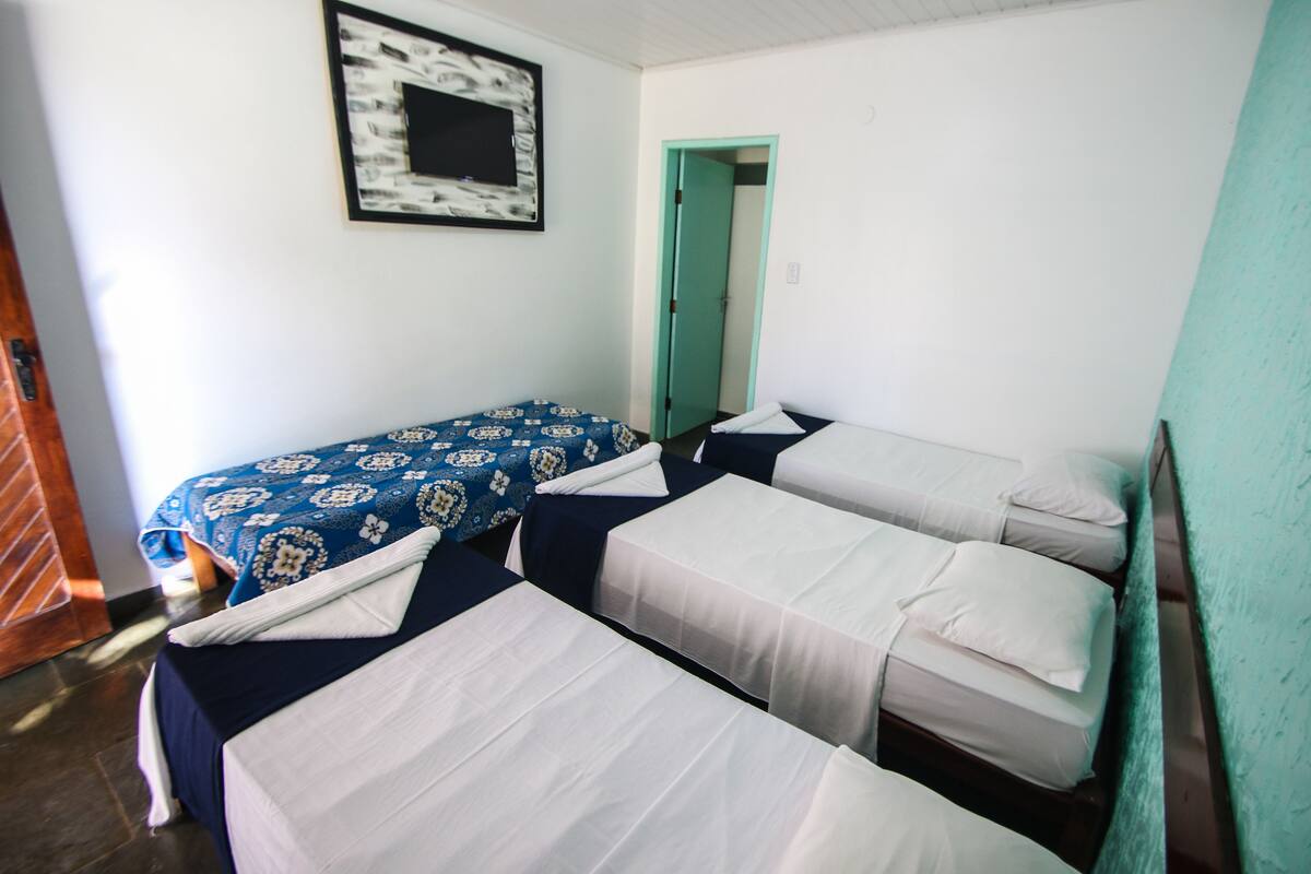 Quarto com duas camas, uma com colcha estampada e outra com roupa de cama lisa. Há um quadro na parede e uma porta com moldura verde.
