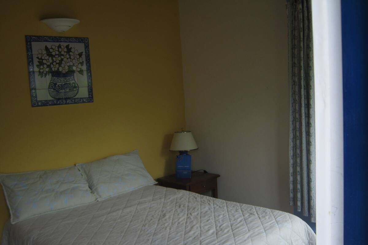Uma cama com cobertura branca acolchoada, uma mesa de cabeceira com abajur e um quadro na parede com um vaso de flores.