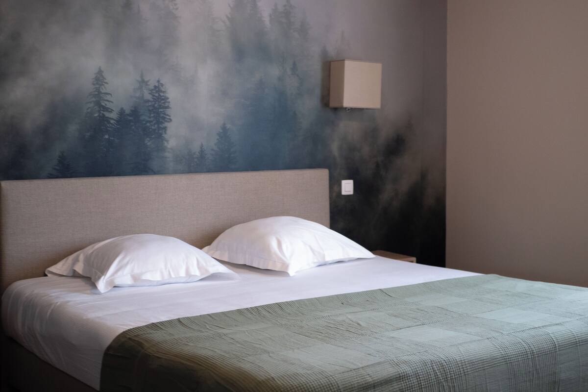 Uma cama com almofadas brancas e um cobertor verde, uma cabeceira e uma parede com um mural de uma floresta.