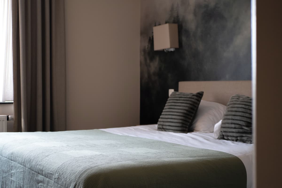 Quarto de hotel com cama, cabeceira, luminária na parede e janela com cortinas.