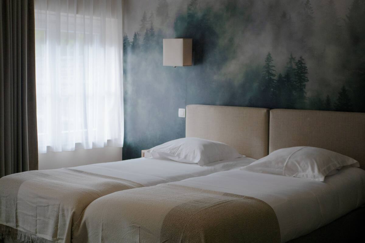 Quarto de hotel com cama, duas almofadas, cabeceira, luminária na parede e janela com cortinas.