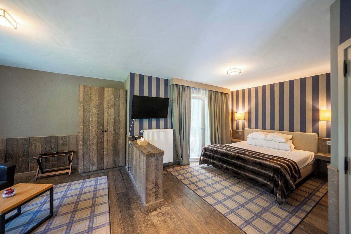 Quarto de hotel com cama, televisão, escrivaninha de madeira, mesa pequena e balcão de recepção.