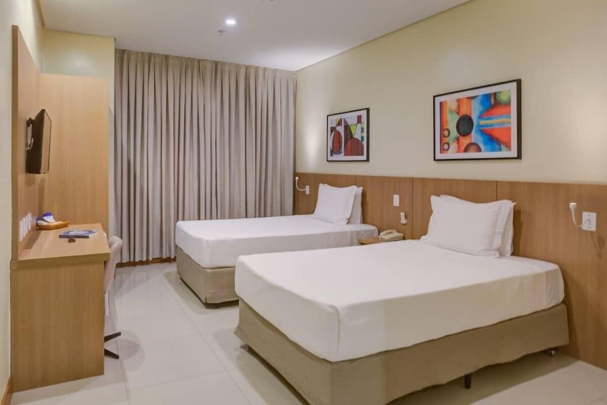 Quarto de hotel com duas camas, uma escrivaninha, um telefone e duas pinturas na parede.