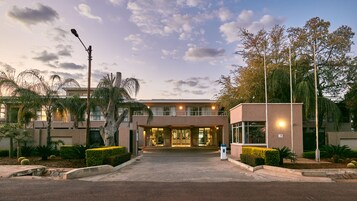 Entrada de hotel com design moderno, apresentando uma estrutura de dois andares, acesso coberto e um jardim com palmeiras e cactos.