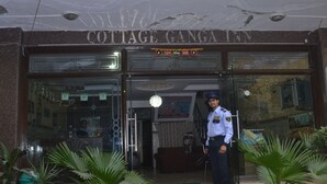 A entrada do Cottage Ganga Inn, com uma recepcionista no interior, um guarda de segurança do lado de fora e plantas em vasos no primeiro plano.