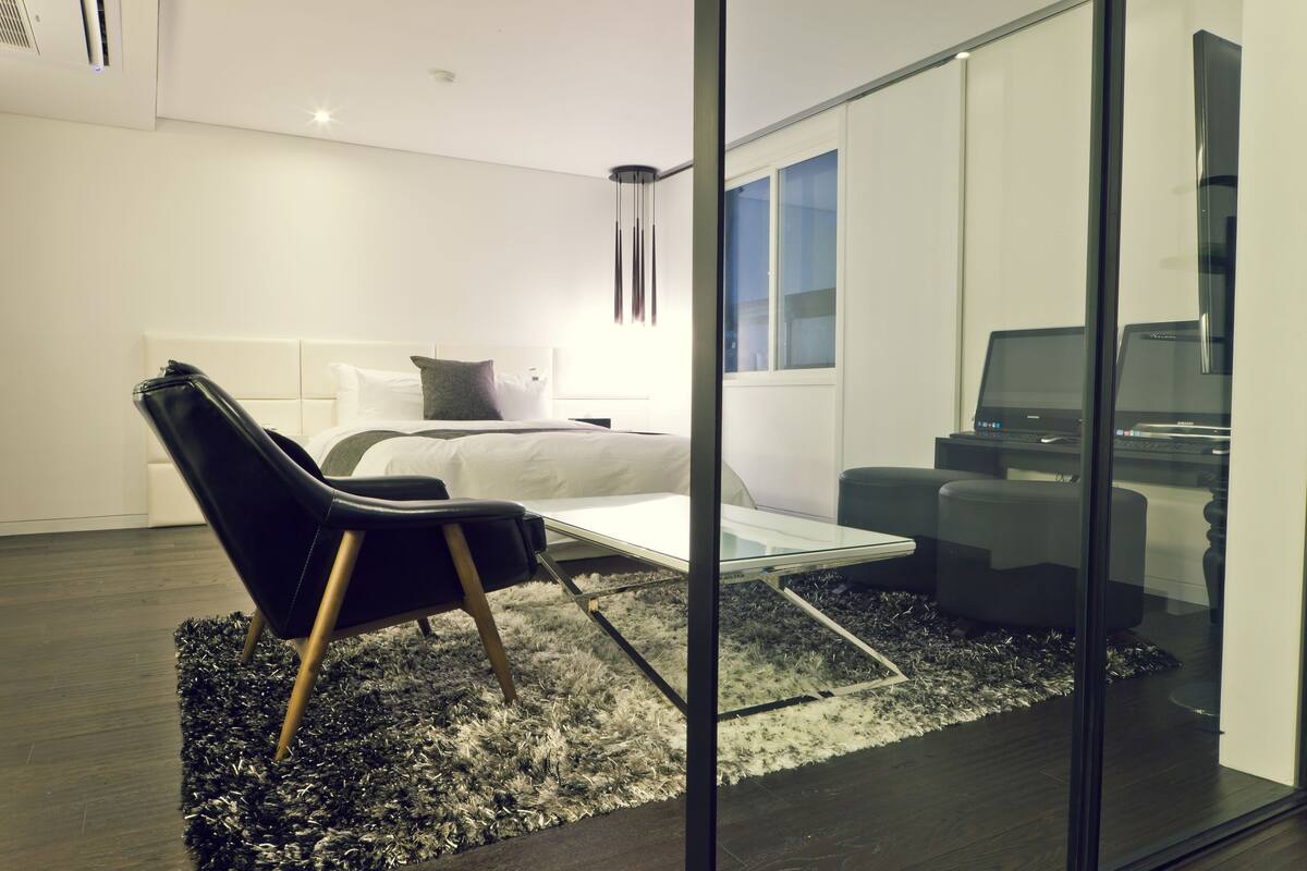 Quarto de hotel moderno com uma cama, uma poltrona de couro preto, uma mesa de centro de vidro e vista para a cidade através de uma janela grande.