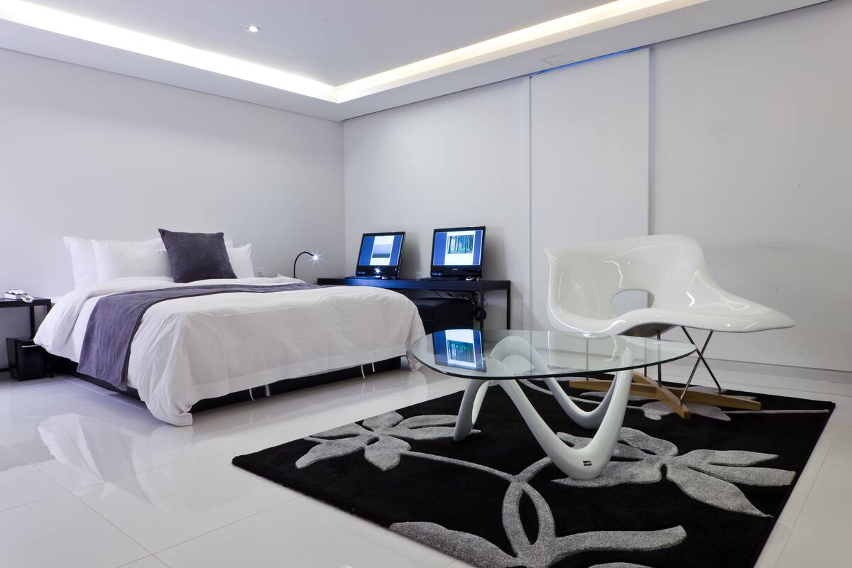 Quarto moderno com uma cama, dois monitores numa escrivaninha e uma mesa de vidro com uma cadeira.