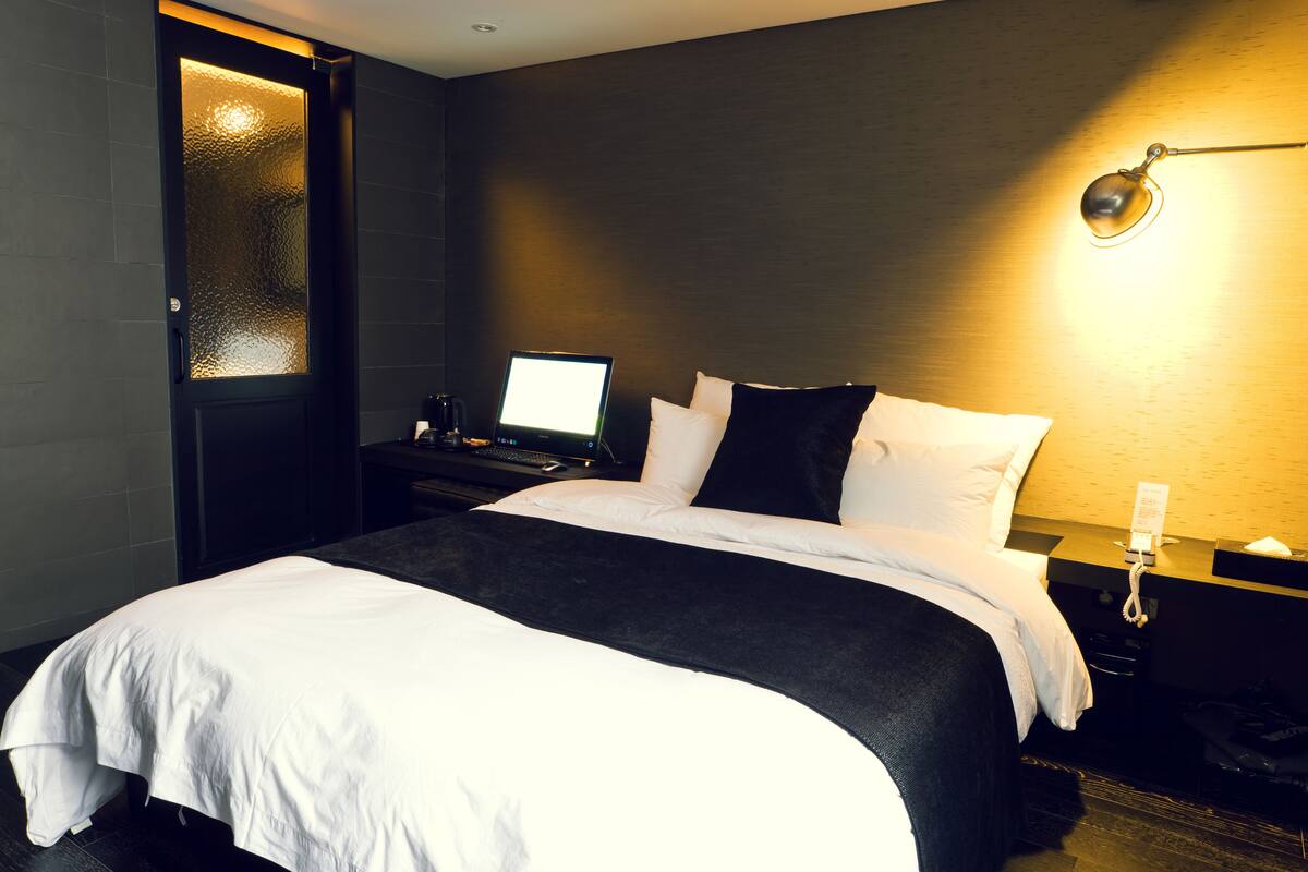 Quarto de hotel com uma cama, uma escrivaninha com computador, uma mesinha de cabeceira e um abajur fixado na parede.