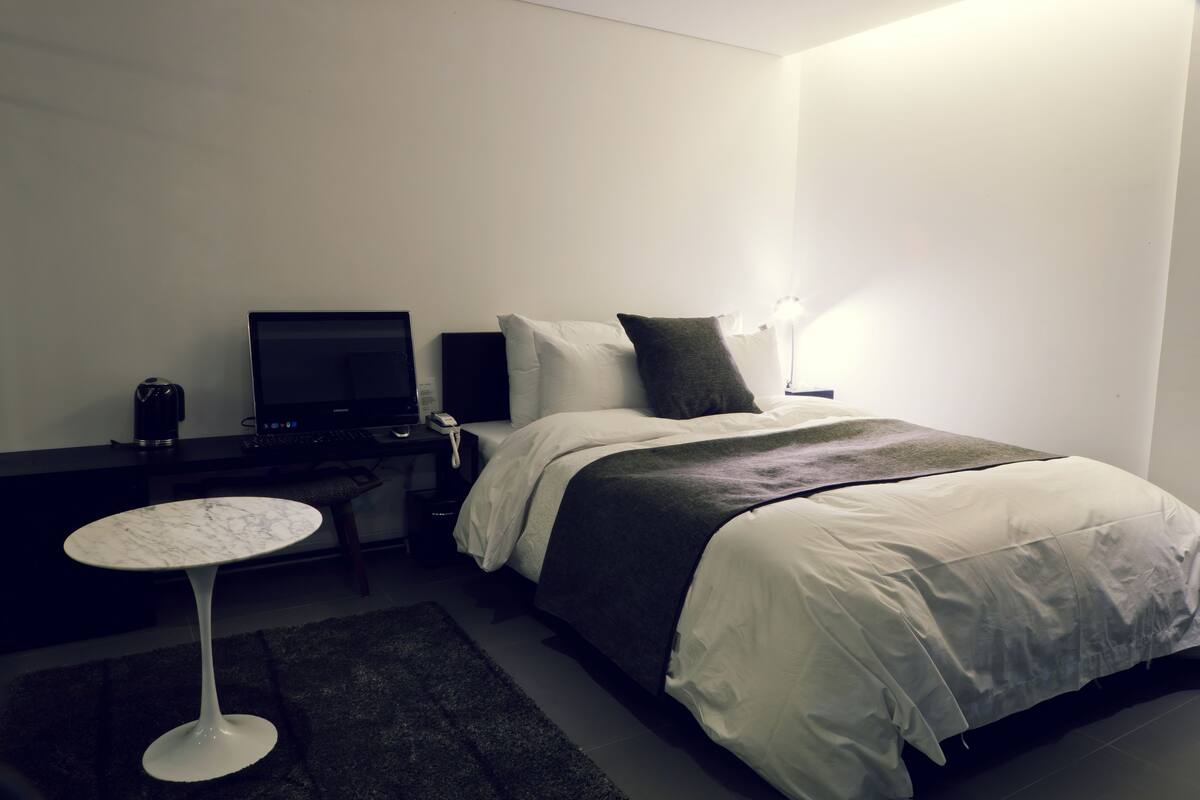 Quarto de hotel moderno com uma cama bem arrumada, uma pequena mesa redonda, uma escrivaninha com um computador e uma televisão, e um abajur.