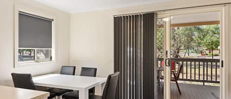 Uma sala de jantar com mesa branca e cadeiras pretas, uma janela com persiana e vista para um deck e vegetação lá fora.