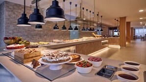 Buffet com diversos doces e bebidas, iluminação pendente moderna e parede de pedra ao fundo.