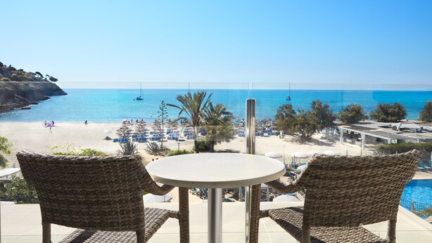 Varanda com cadeiras de vime e mesa, com vista para uma praia com guarda-sóis e piscina.