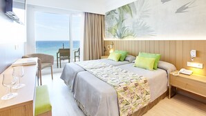 Quarto de hotel com cama, mesa de cabeceira e escrivaninha com dois copos. Vista para o oceano através de uma grande janela.
