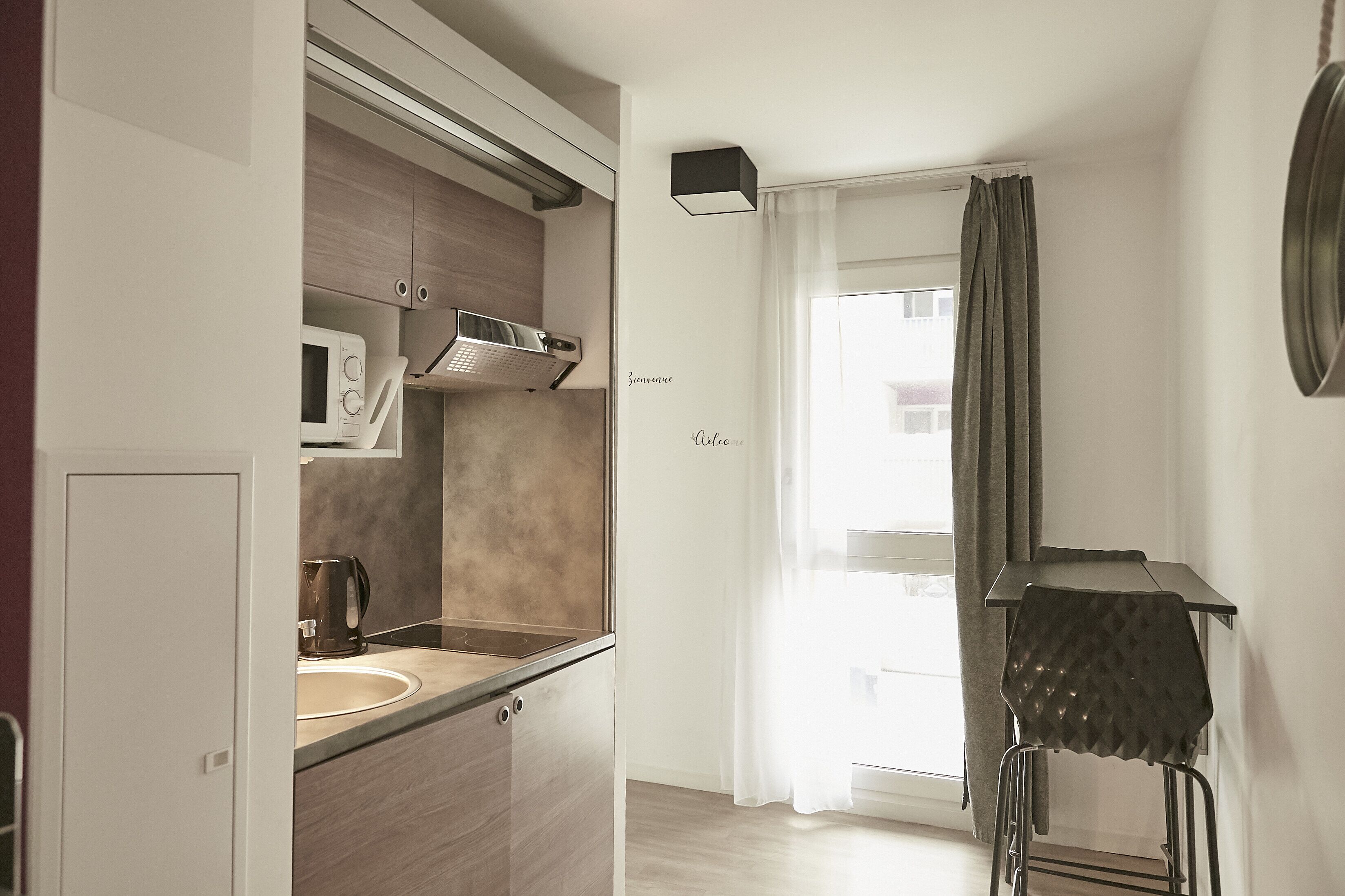 Cozinha moderna com micro-ondas, chaleira e forno embutido. Há uma janela com cortinas e uma pequena mesa com uma cadeira.