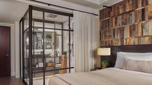 Quarto de hotel com uma cama grande, área de chuveiro com divisória de vidro, painel de parede de madeira e mesa de cabeceira com abajur.