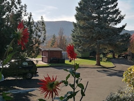 Uma entrada de casa com uma pequena cabana de madeira, um carro estacionado e uma montanha ao fundo.