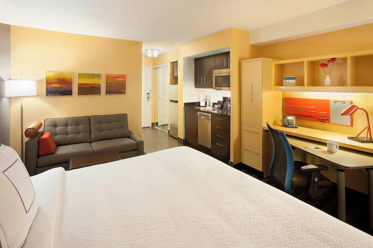 Quarto de hotel com cama, sofá, escrivaninha, cadeira, luminária e cozinha equipada com armários e eletrodomésticos.