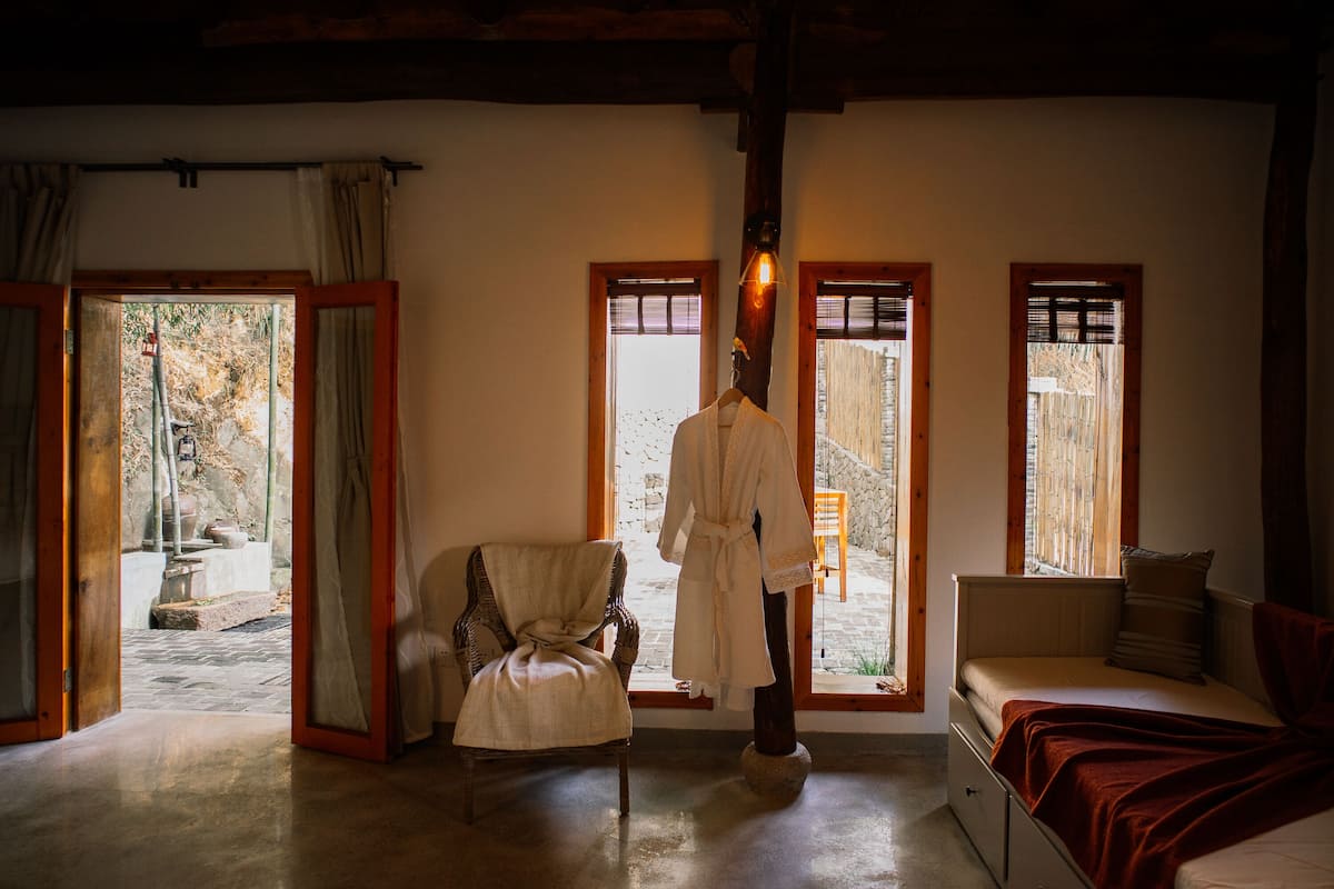 Quarto com cama, uma cadeira e um roupão pendurado em um gancho. Há janelas com molduras de madeira e uma parede de pedra visível através delas.