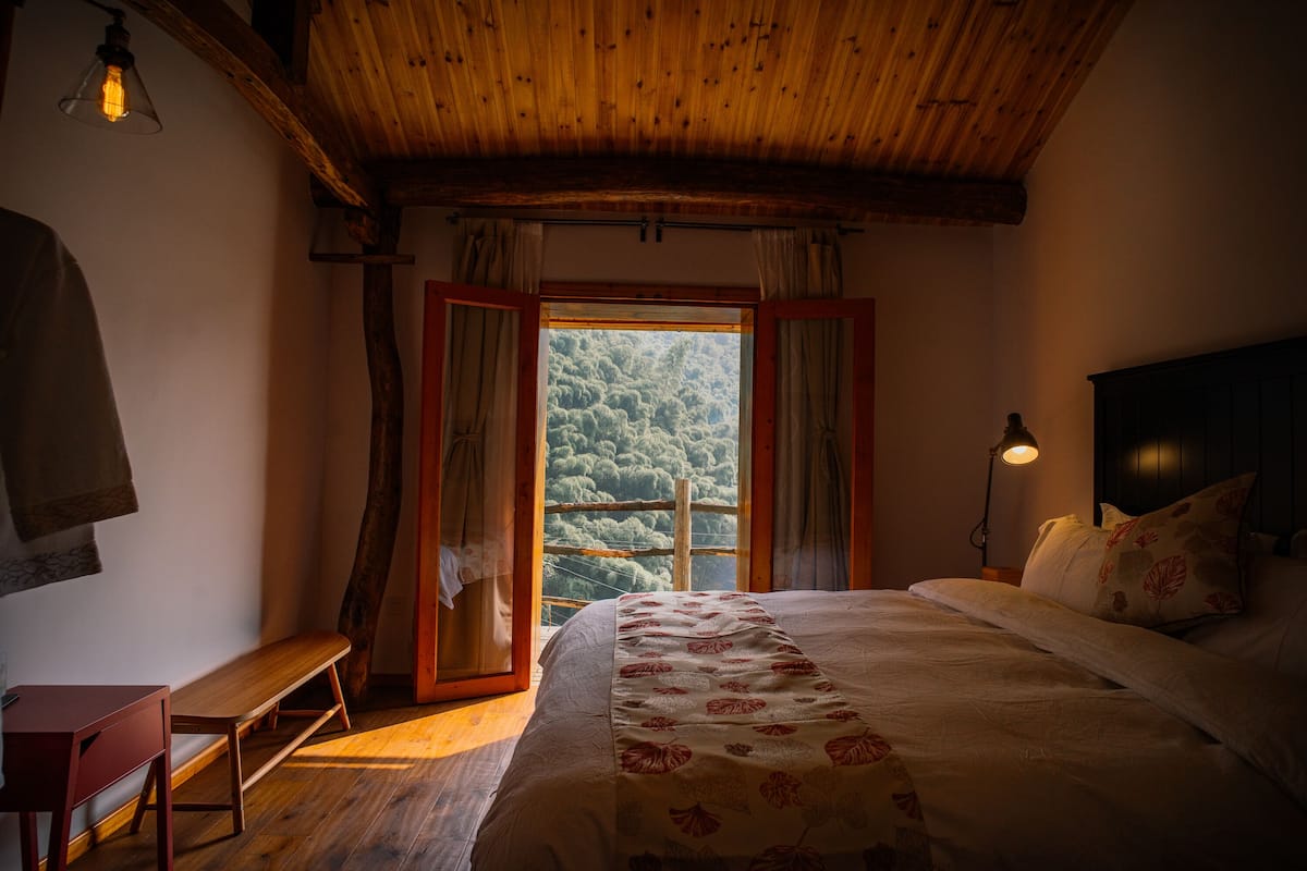 Quarto com teto de madeira, cama grande com roupa de cama floral, um banco e vista para árvores através de uma porta aberta.