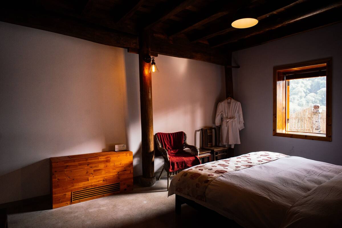 Um quarto com uma cama, um banco de madeira, uma cadeira com um cobertor vermelho, uma mesa de madeira e uma janela com vista para árvores.