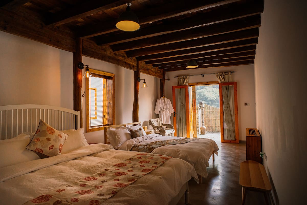 Quarto com duas camas, vigas de madeira, luminária pendente e vista por uma porta.