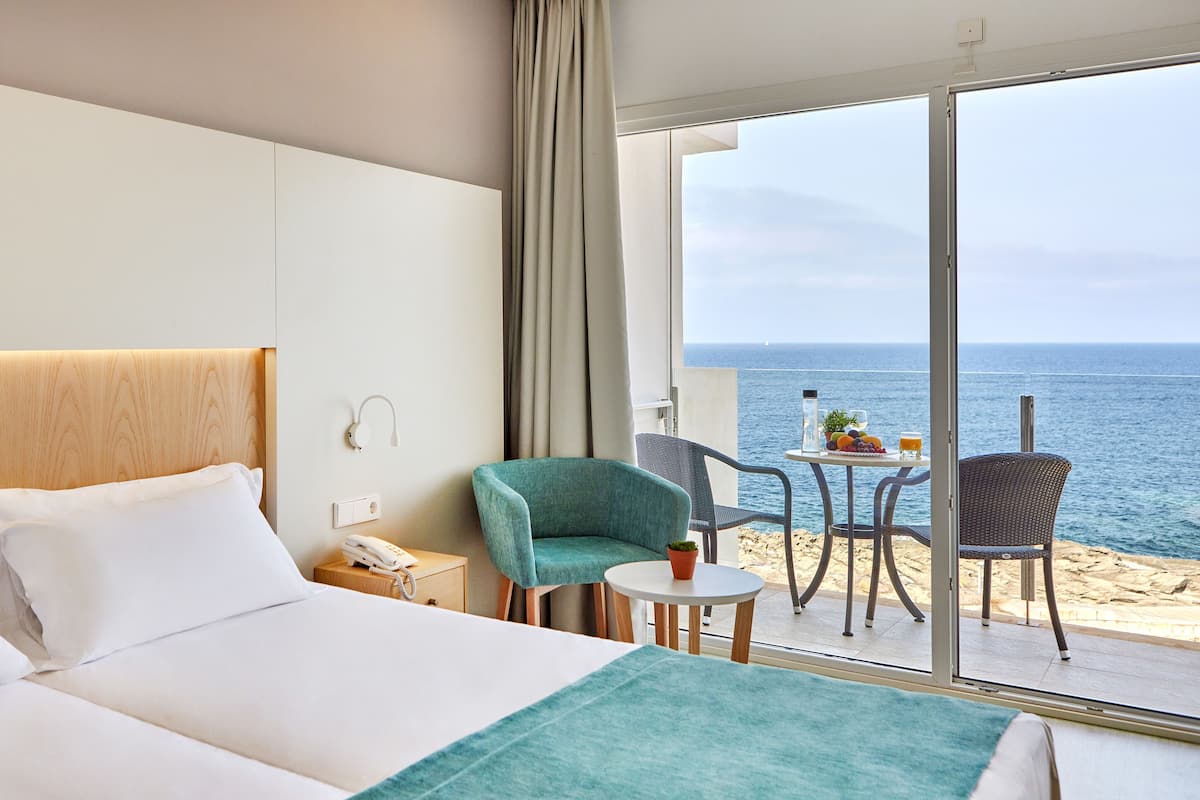 Quarto de hotel com cama, uma mesinha com uma travessa de frutas e bebidas, uma cadeira e vista para o mar.