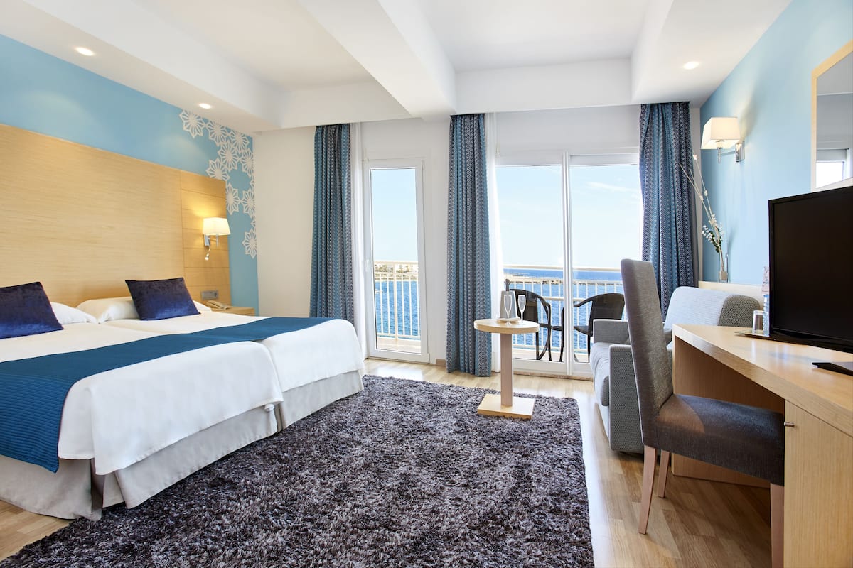 Quarto de hotel com duas camas, uma escrivaninha, uma cadeira, televisão e varanda com vista.
