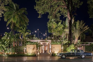 Entrada de hotel com um carro estacionado na frente, cercada por vegetação tropical e iluminada por luzes de cordão.