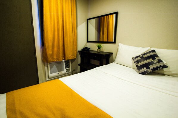 Quarto de hotel com uma cama, mesa de cabeceira com telefone, espelho e janela com cortina.