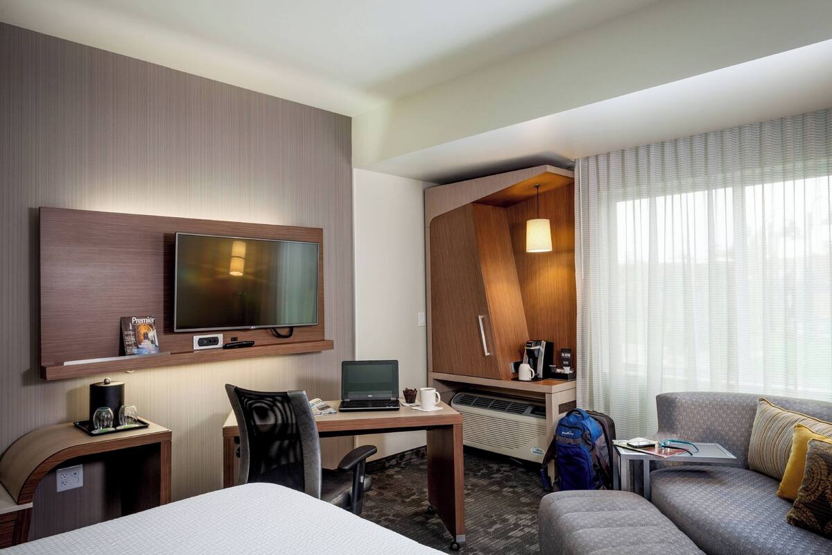 Quarto de hotel com cama, escrivaninha com laptop, televisão, cafeteira e sofá.