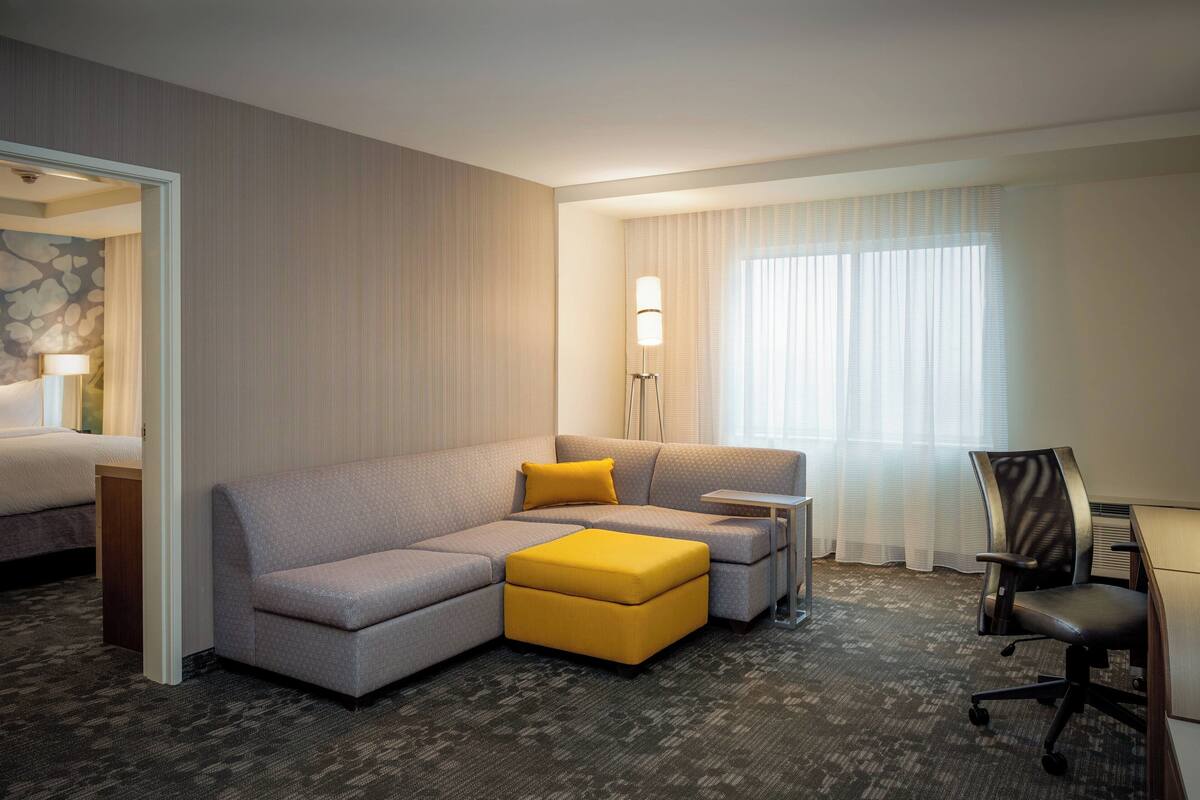Quarto de hotel com sofá de canto, um pequeno banquinho, escrivaninha com cadeira e uma janela grande com cortinas.