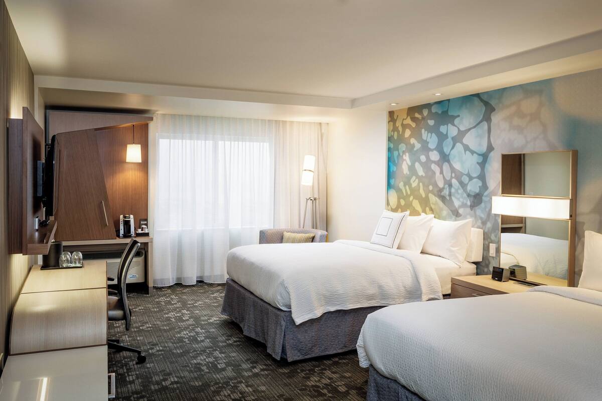Quarto de hotel com duas camas, uma escrivaninha, uma cadeira, uma televisão e uma janela grande com cortinas.