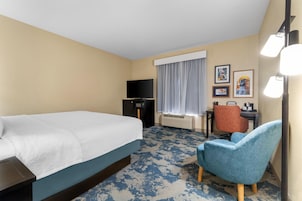 Quarto de hotel com cama, escrivaninha e cadeira, televisão, janela com cortinas e obras de arte na parede.