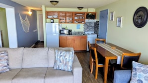 Uma sala de estar com sofá, uma mesa de jantar com cadeiras, uma cozinha com armários e eletrodomésticos, e um relógio na parede.