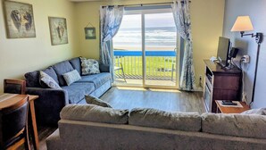 Uma sala de estar com sofá, televisão e vista para o oceano através de uma porta de vidro deslizante.