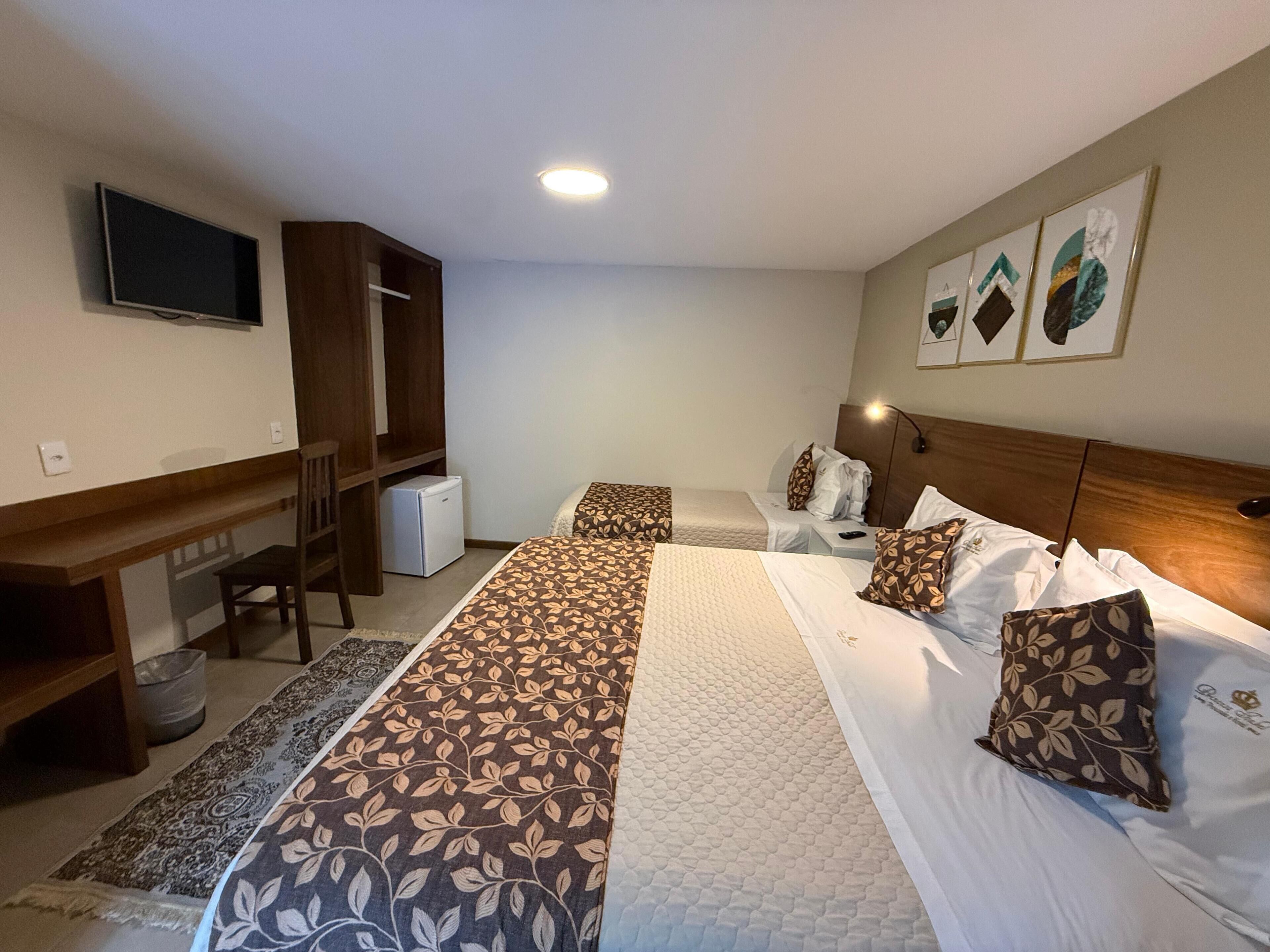 Quarto de hotel com cama, escrivaninha, cadeira, televisão e um pequeno frigorífico.