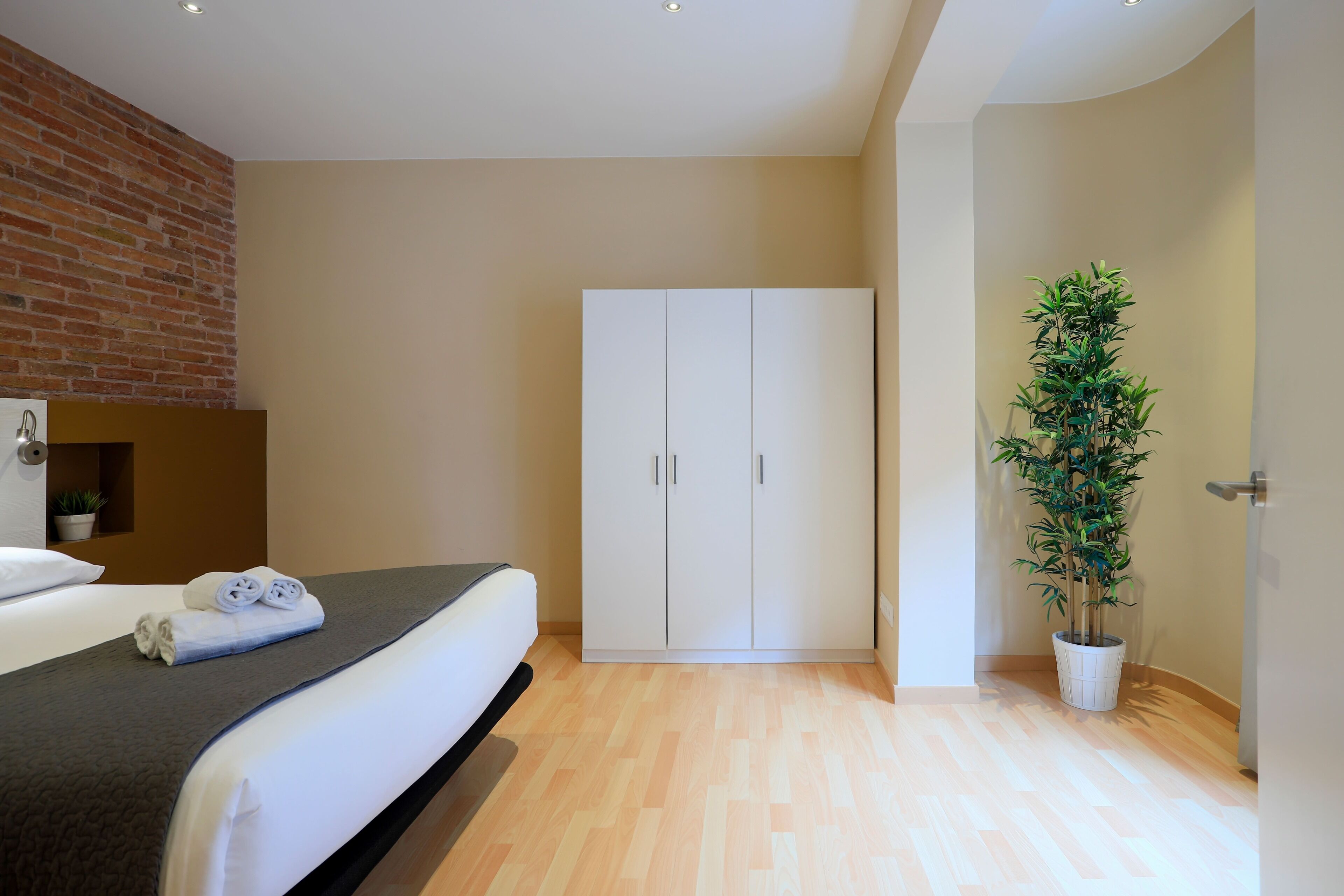 Quarto de hotel moderno com cama, guarda-roupa, planta em vaso e parede de tijolos.