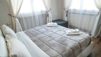 Uma cama bem arrumada com colcha cinza, uma pequena mesa de cabeceira com toalha dobrada e cortinas finas numa janela.