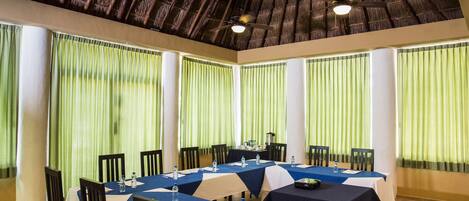 Uma sala de conferências com mesas preparadas para reuniões, janelas amplas com cortinas verdes e teto de palha.