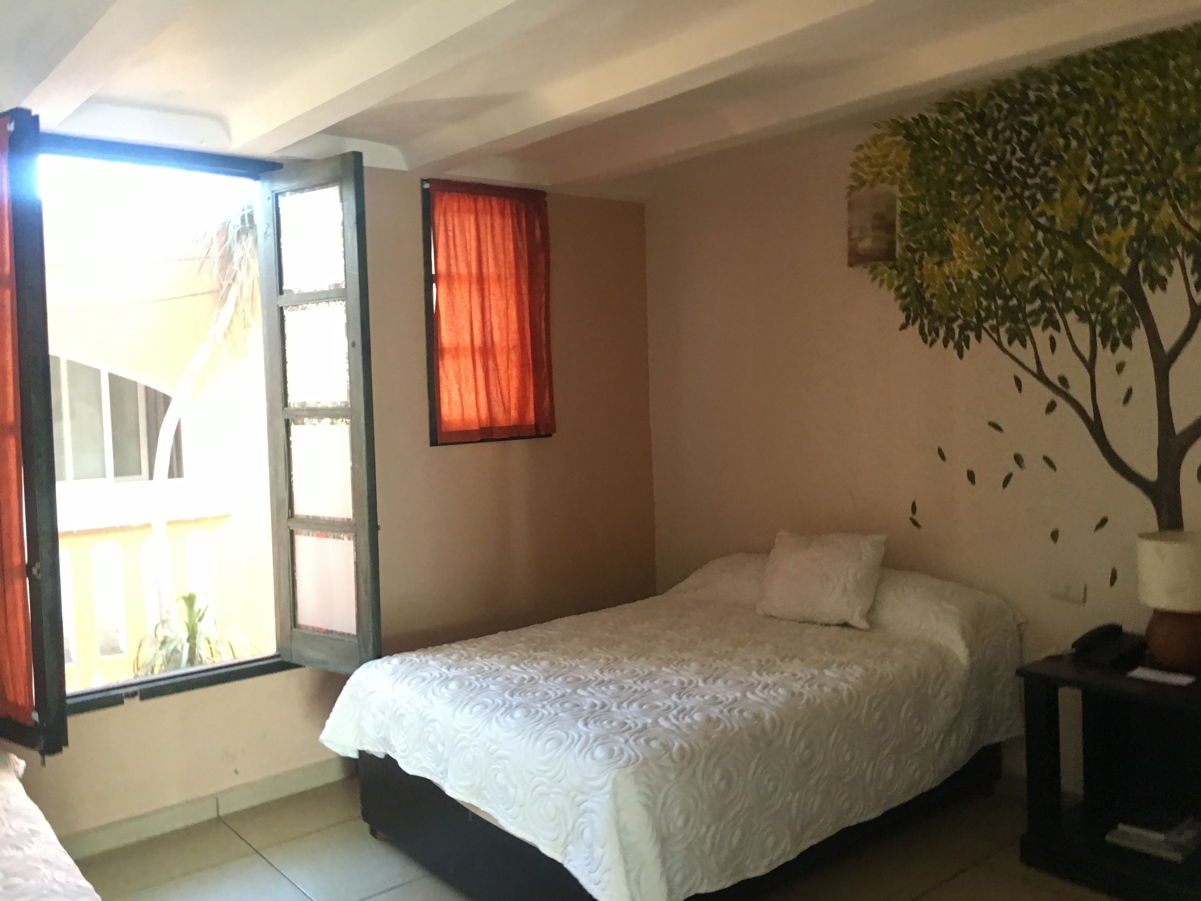 Quarto com cama, mesa de cabeceira, janela com cortinas e um mural de árvore na parede.