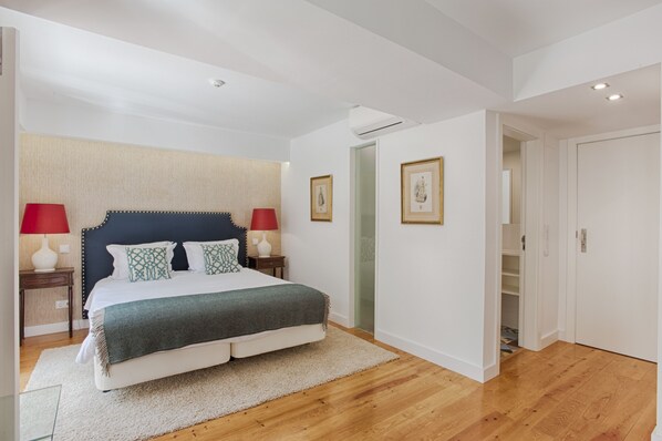 Quarto com cama, duas mesas de cabeceira com abajures, piso de madeira e porta que dá para um armário.