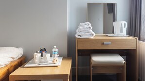 Quarto de hotel com cama, escrivaninha de madeira com espelho, pequena mesa com bandeja de produtos de higiene e janela com cortinas.