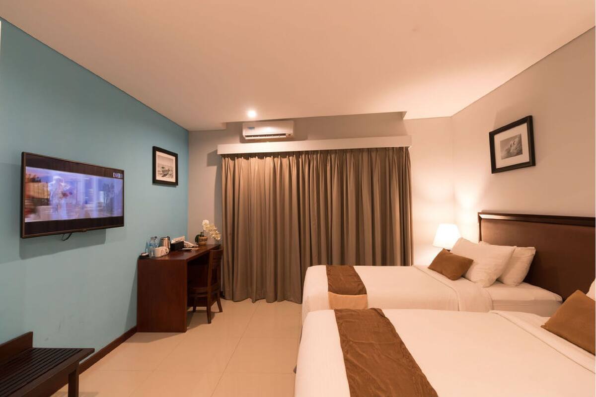 Quarto de hotel com duas camas, uma escrivaninha, televisão e janela com cortinas.